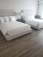 Quarto de hotel com duas camas, uma mesa de cabeceira com abajur e uma cadeira.