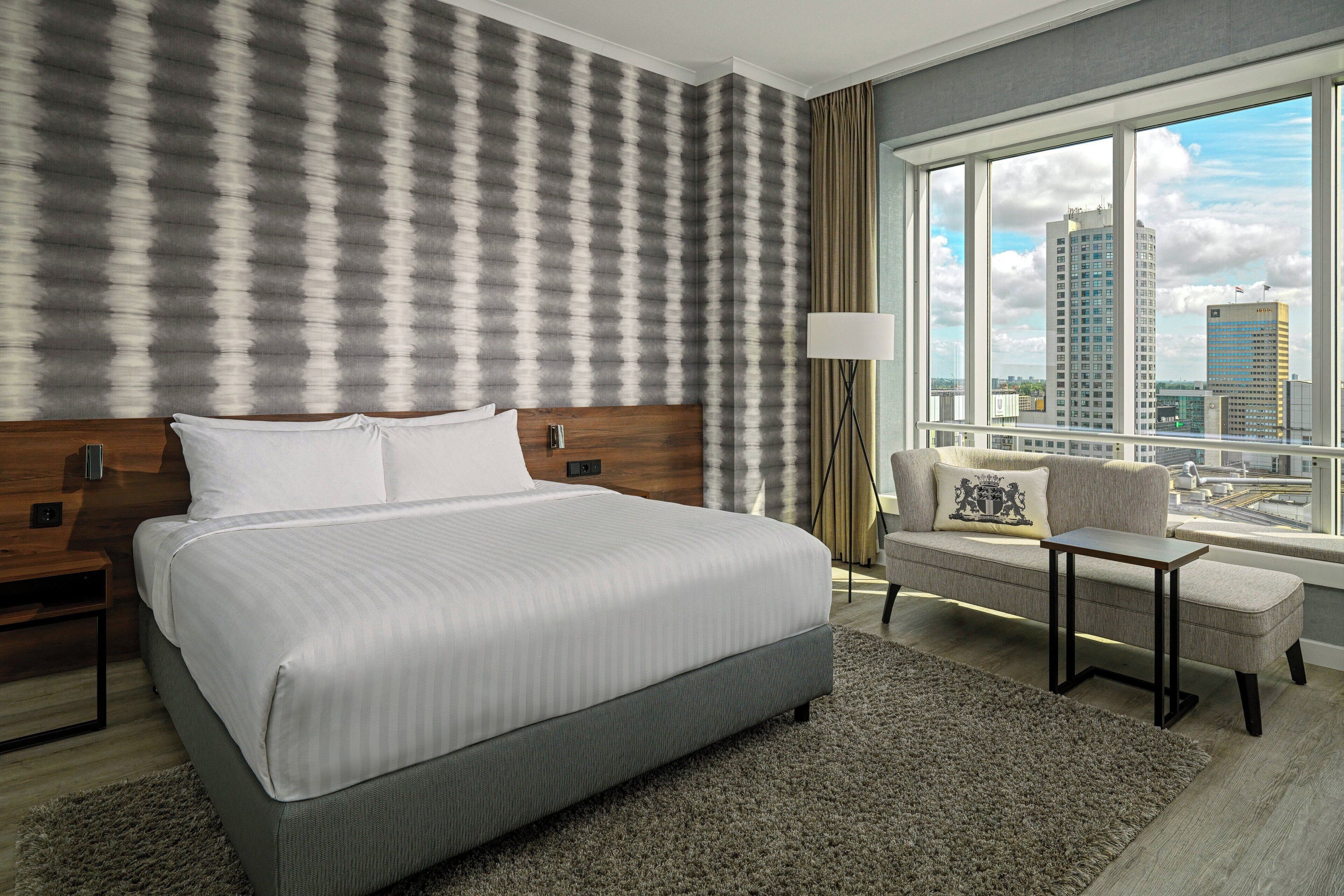 Quarto de hotel moderno com uma cama grande, um sofá, uma mesinha e vista para a paisagem urbana através de grandes janelas.