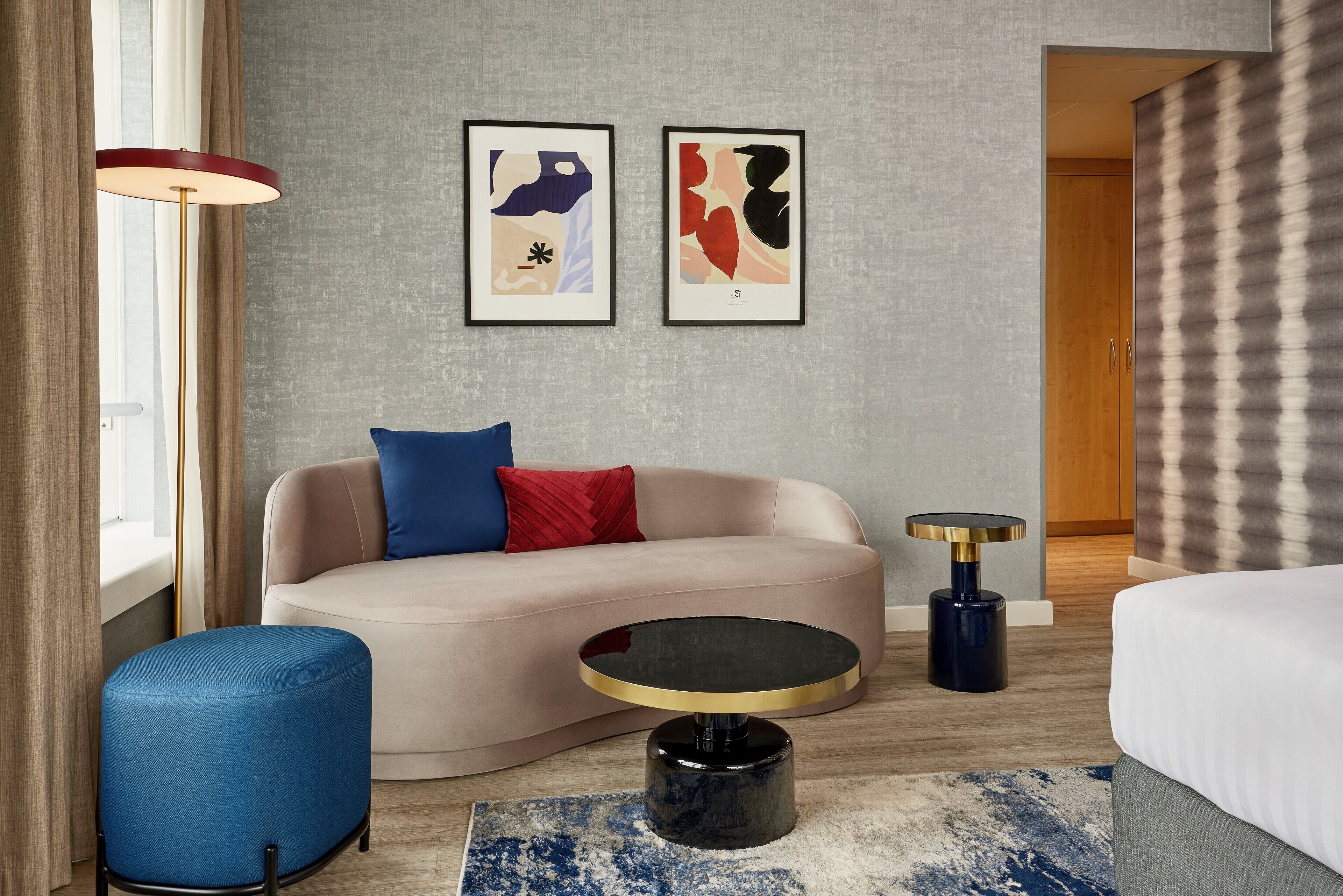 Quarto de hotel moderno com sofá curvo, mesa de centro redonda, um banquinho azul e uma cama. Há duas obras de arte emolduradas na parede e uma cortina estampada na janela.