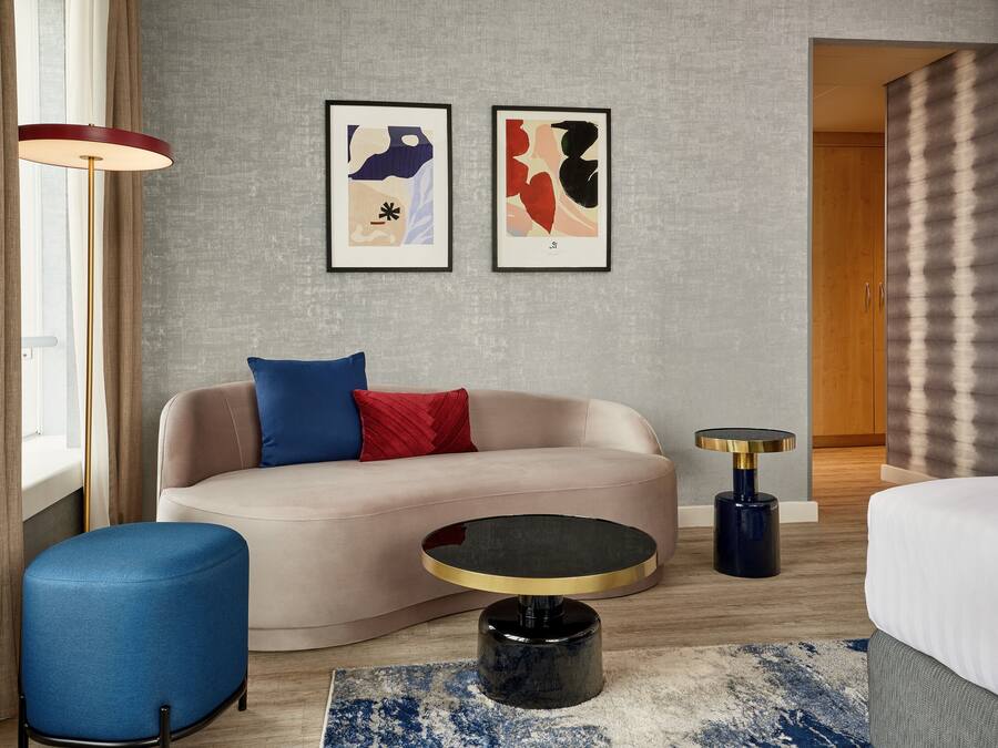 Quarto de hotel moderno com sofá curvo, mesa de centro redonda, um banquinho azul e uma cama. Há duas obras de arte emolduradas na parede e uma cortina estampada na janela.
