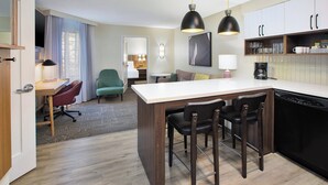 Quarto de hotel moderno com cozinha, banquetas de bar, mesa de jantar e uma poltrona verde.