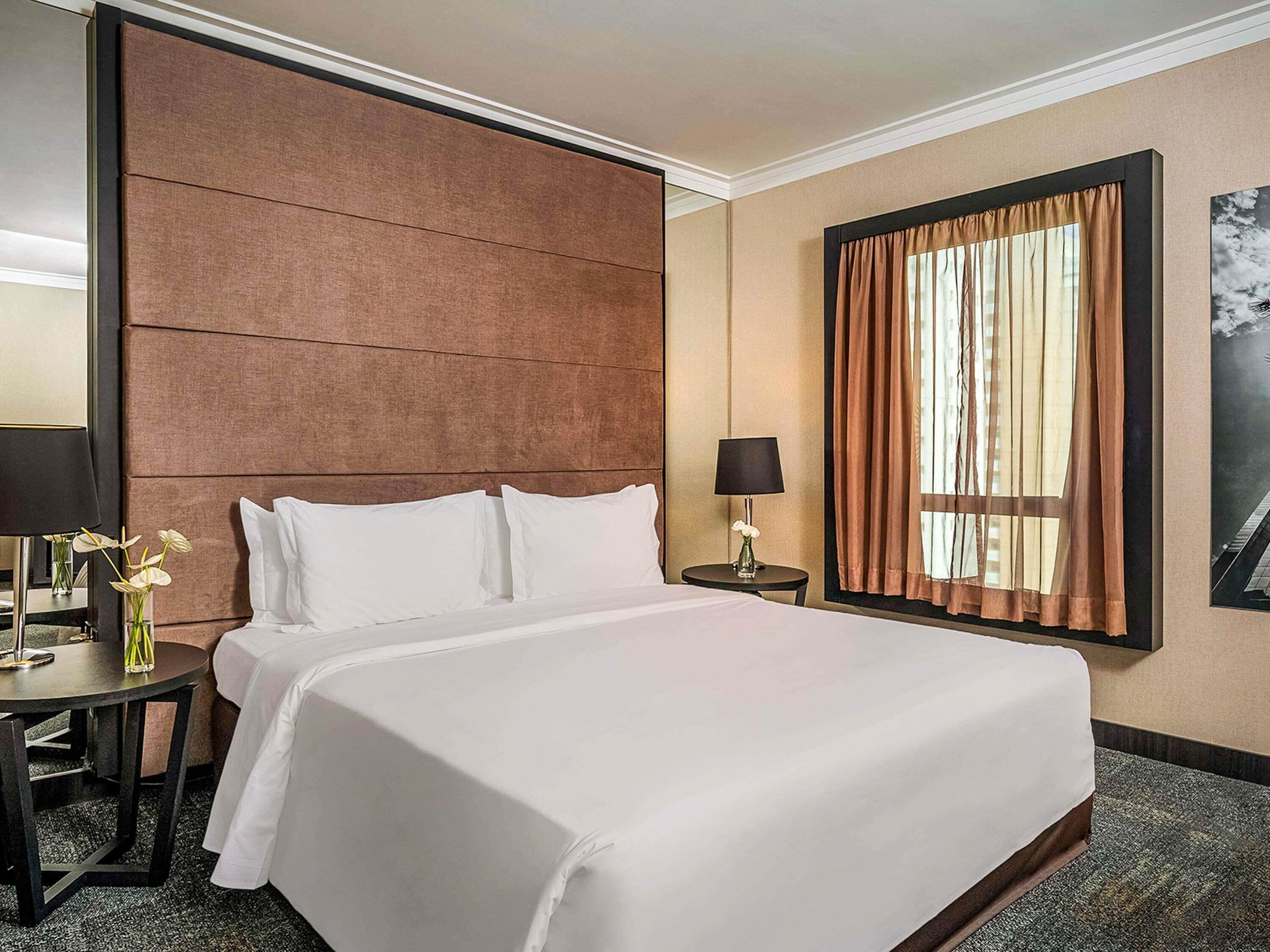 Quarto de hotel com uma cama grande, uma mesa de cabeceira com abajur, uma pequena mesa com um vaso de flores e uma janela com cortinas.