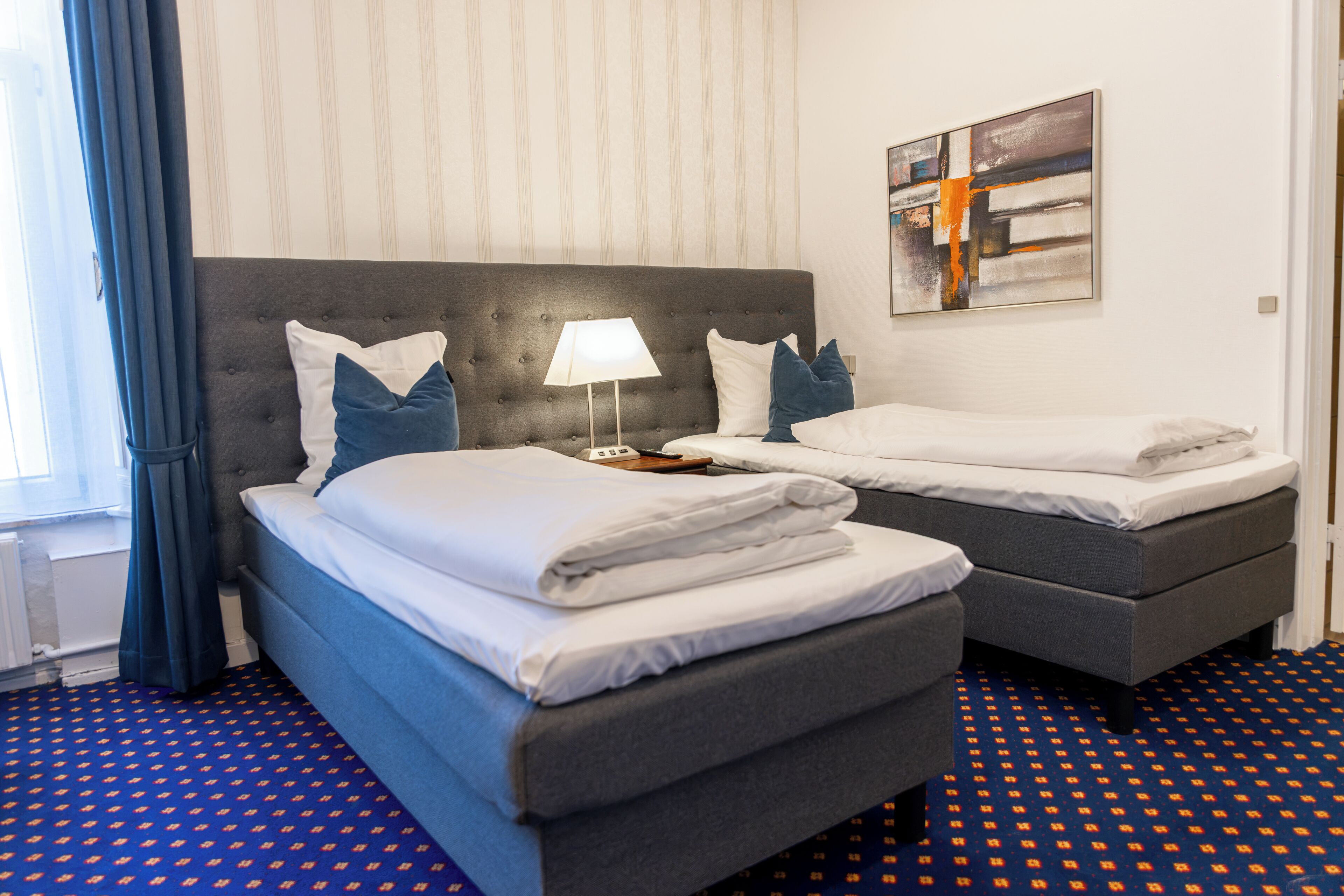 Quarto de hotel com uma cama grande, uma cama menor, uma mesinha de cabeceira, uma luminária e um quadro na parede.