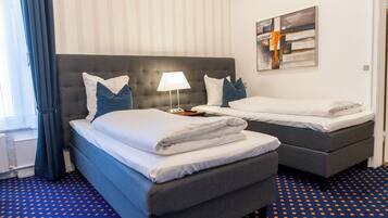 Quarto de hotel com uma cama grande, uma cama menor, uma mesinha de cabeceira, uma luminária e um quadro na parede.
