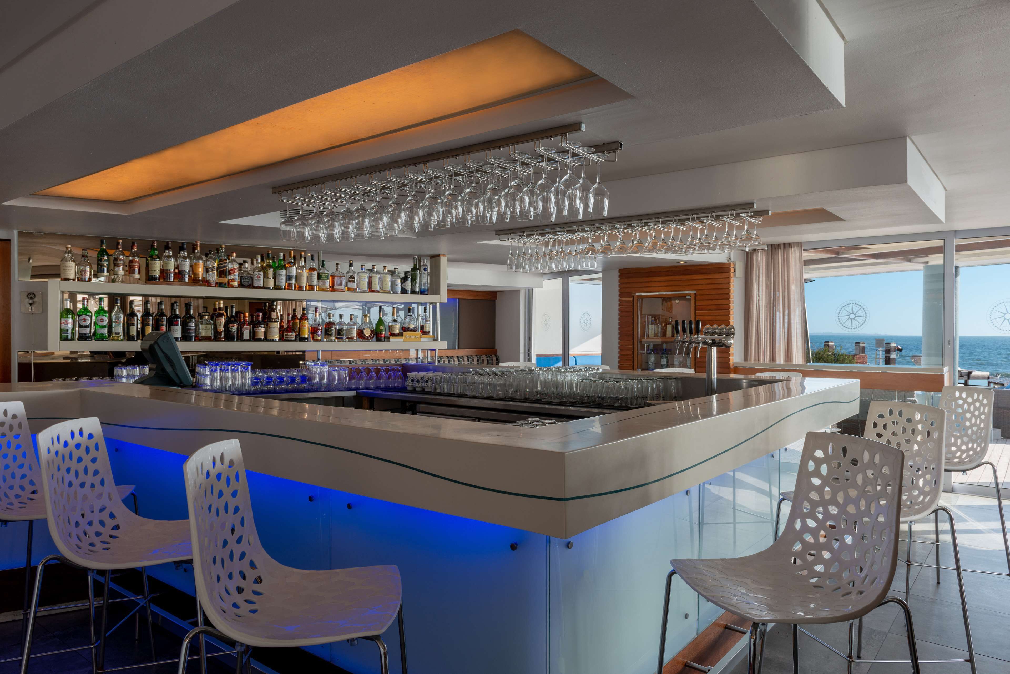 Uma área de bar moderna com balcão de vidro, diversas opções de assento e vista para o mar.
