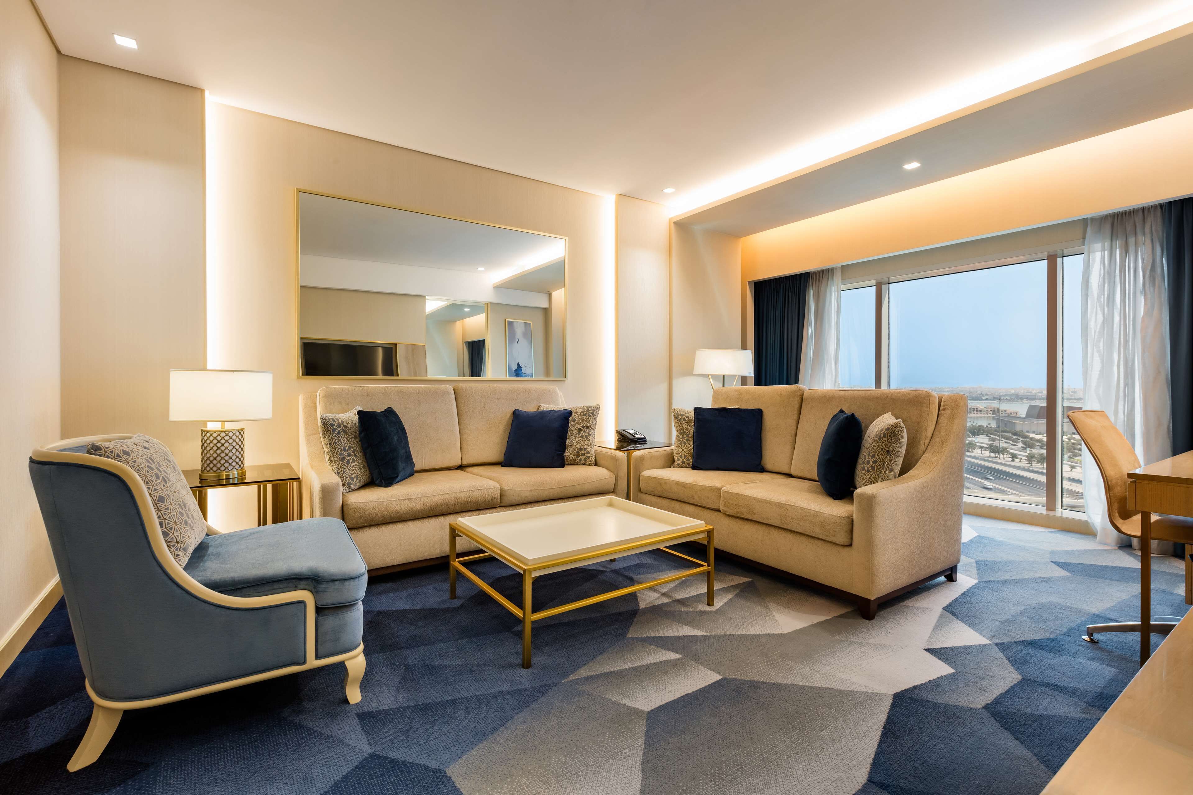 Quarto de hotel moderno com um amplo sofá, uma mesa de centro e vista para a cidade através de grandes janelas.