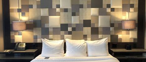 Quarto de hotel com design moderno, com uma cama grande, roupa de cama branca, duas mesas de cabeceira com luminárias e uma parede com padrão geométrico.