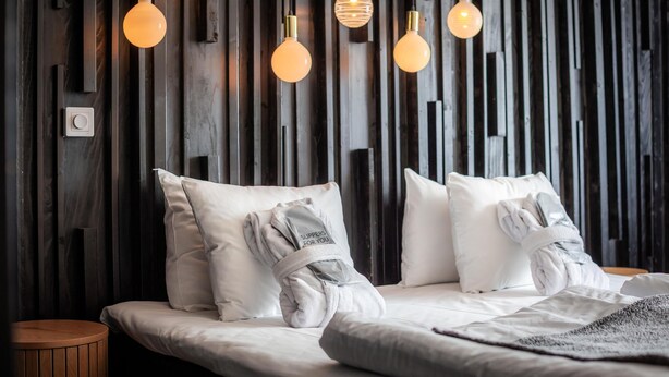 Uma cama com roupa de cama branca, parede de madeira e luminárias pendentes.