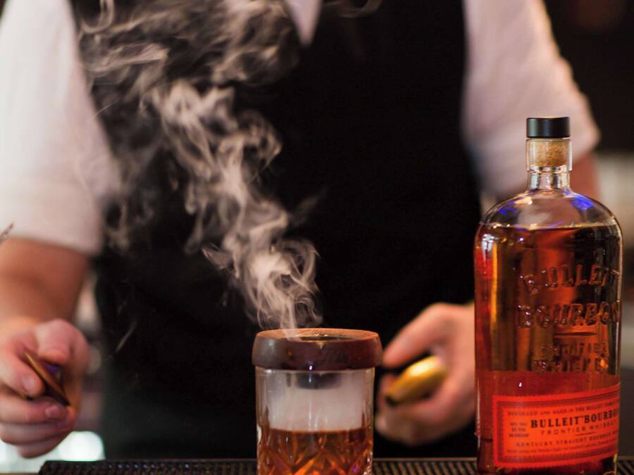 Um bar com uma garrafa de Bulleit Bourbon, um copo com bebida e alguém servindo.