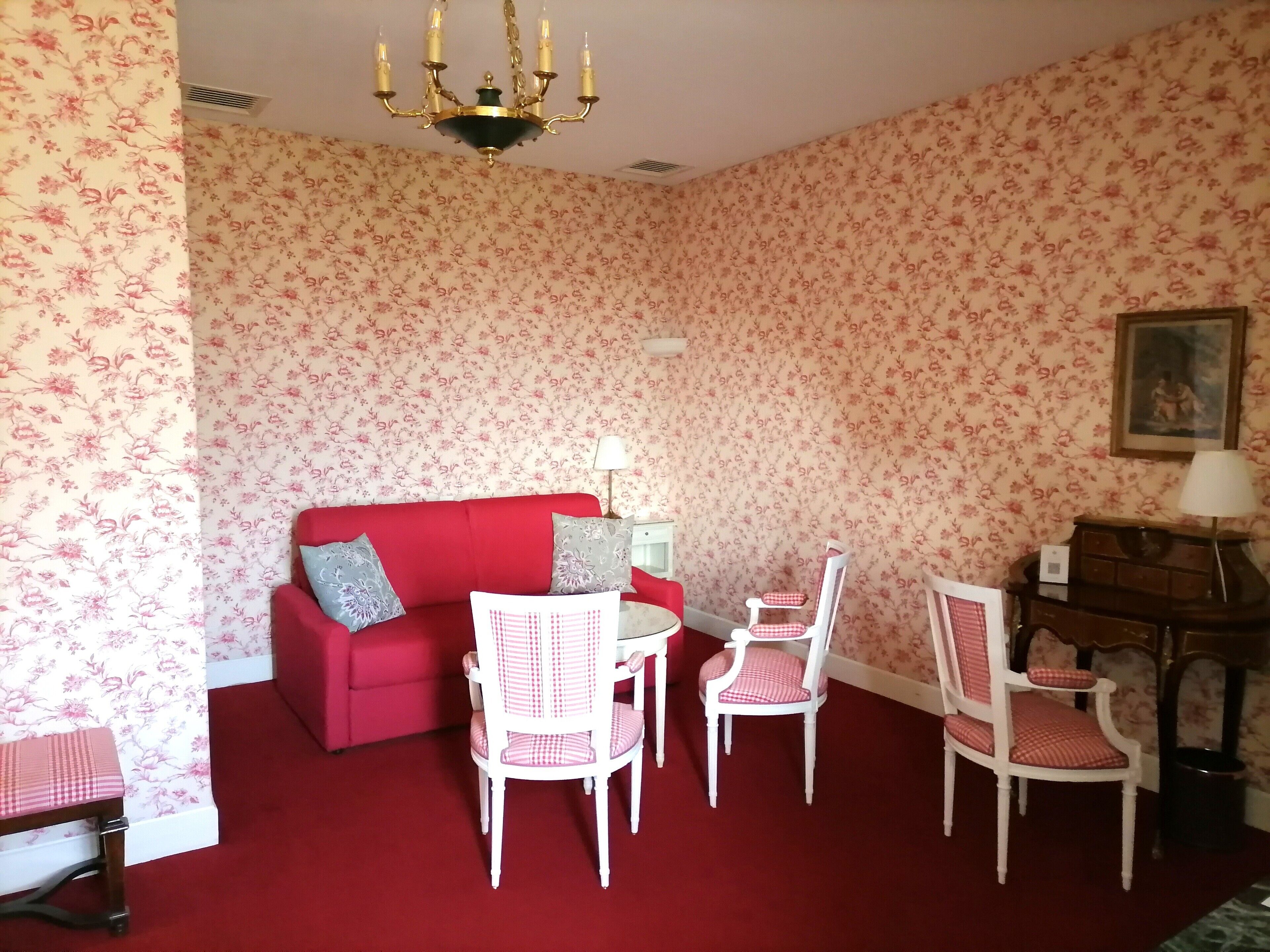 Quarto com papel de parede floral, um sofá vermelho, uma mesa pequena com cadeiras e um candelabro.