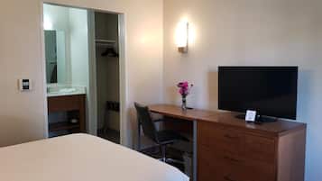 Quarto de hotel com cama, escrivaninha com cadeira, televisão, guarda-roupa, espelho e luminária na parede.