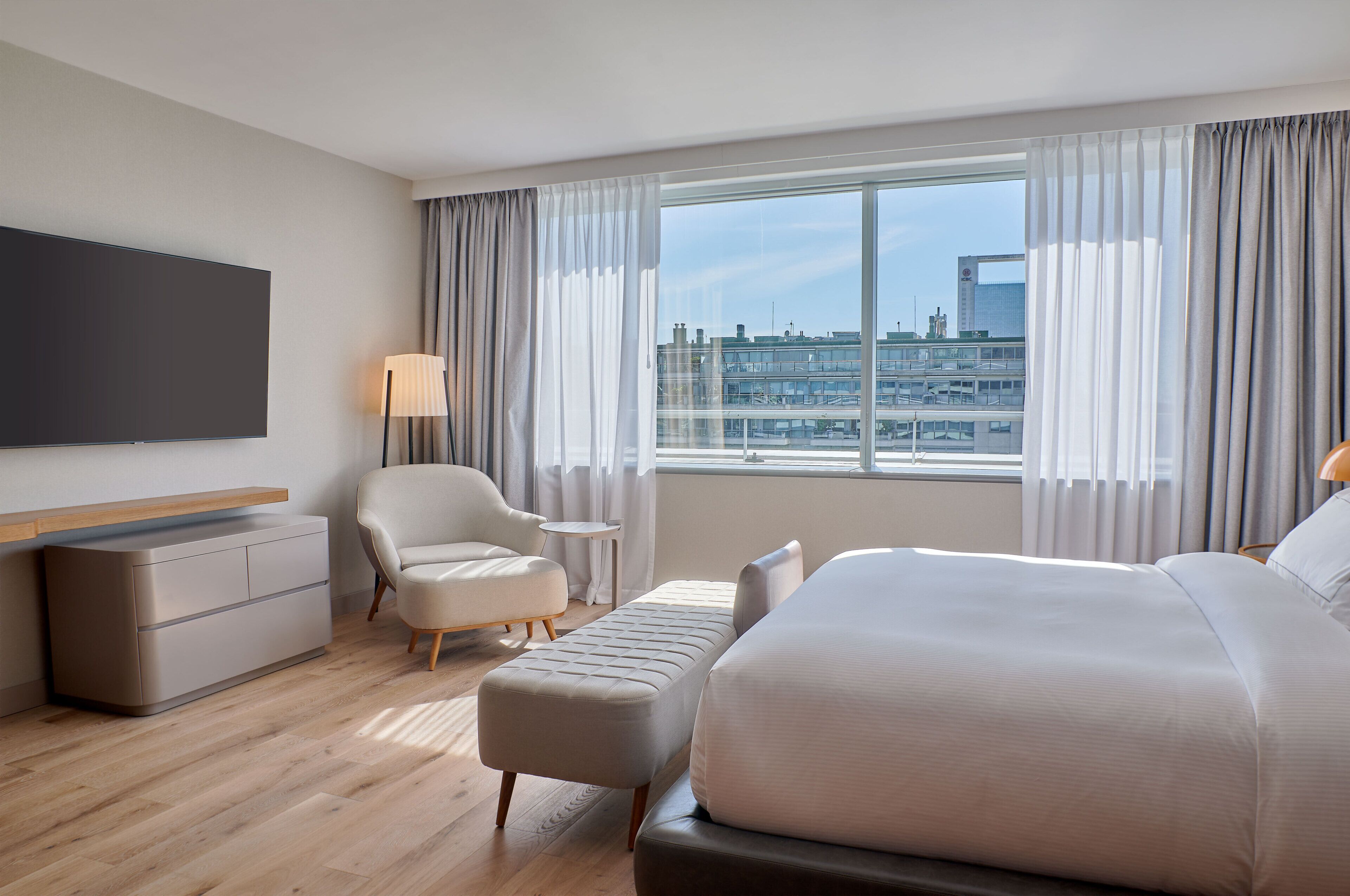 Quarto de hotel moderno com uma cama grande, um sofá, uma TV, uma mesa pequena e vista para a cidade através de grandes janelas.