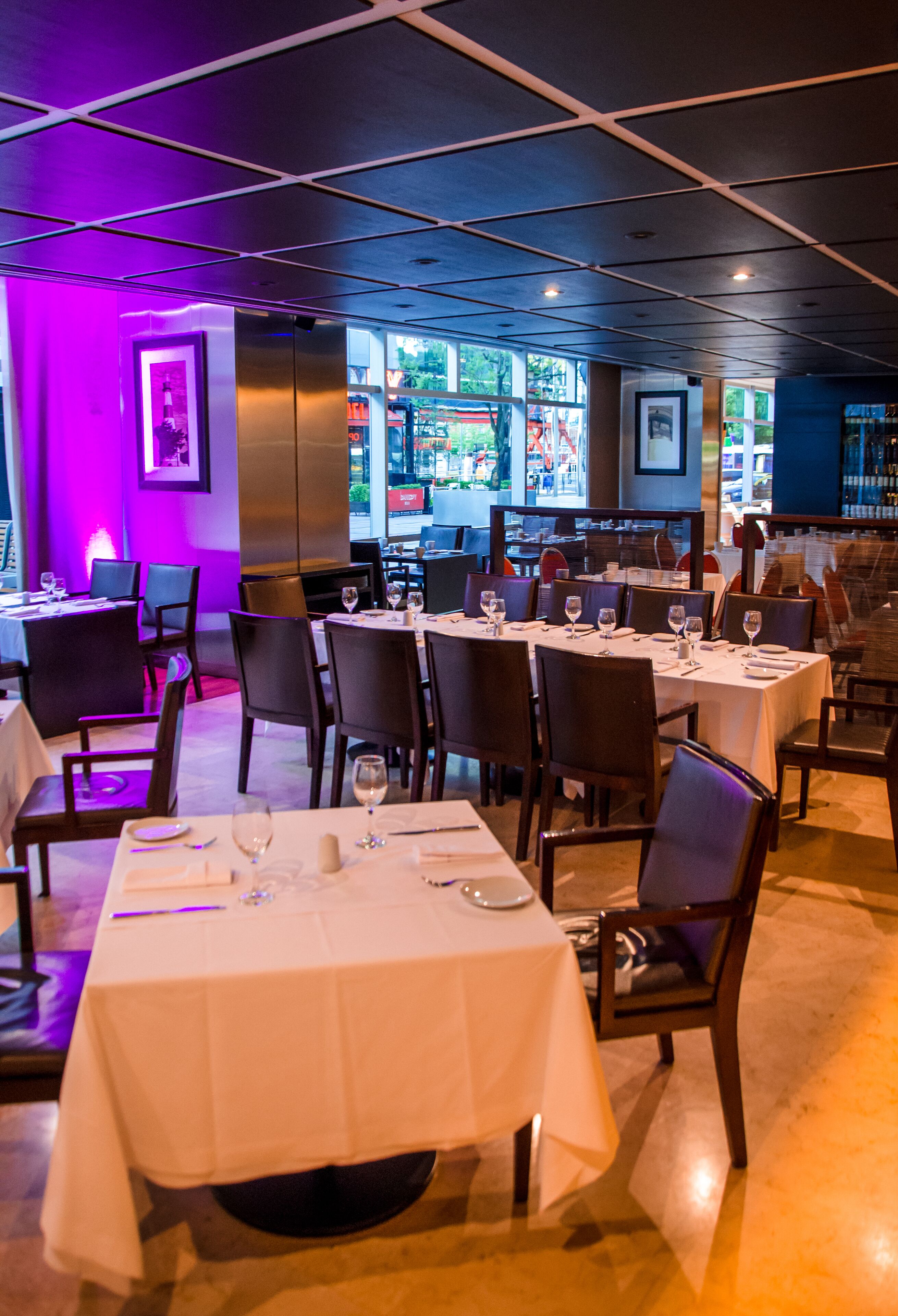 Restaurante com mesas postas para refeições, área de bar e vista para a paisagem urbana através de grandes janelas.