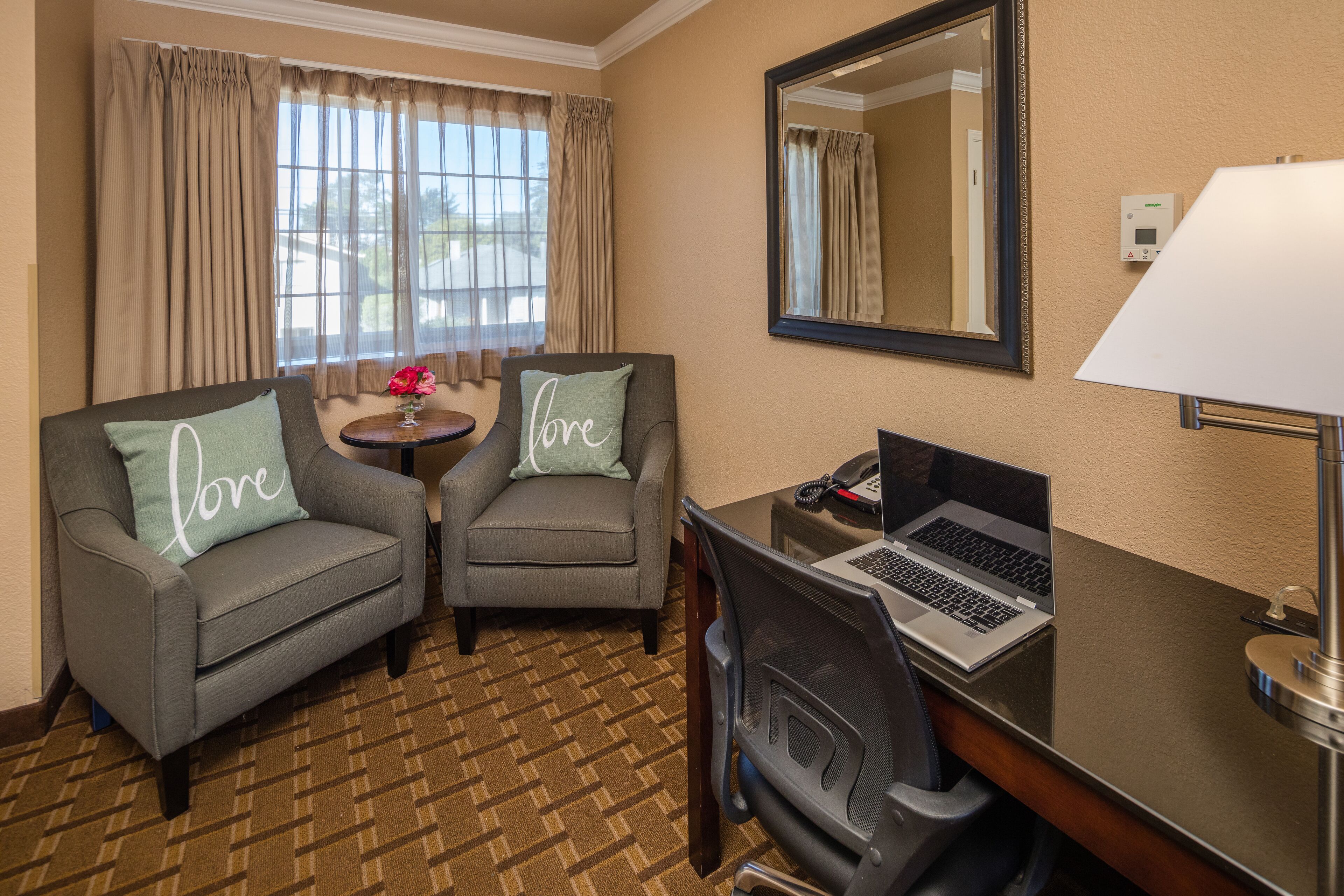 Quarto de hotel com duas poltronas, uma mesinha de centro com um vaso de flores, uma escrivaninha com um laptop, uma luminária e um espelho grande na parede.