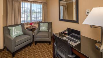 Quarto de hotel com duas poltronas, uma mesinha de centro com um vaso de flores, uma escrivaninha com um laptop, uma luminária e um espelho grande na parede.