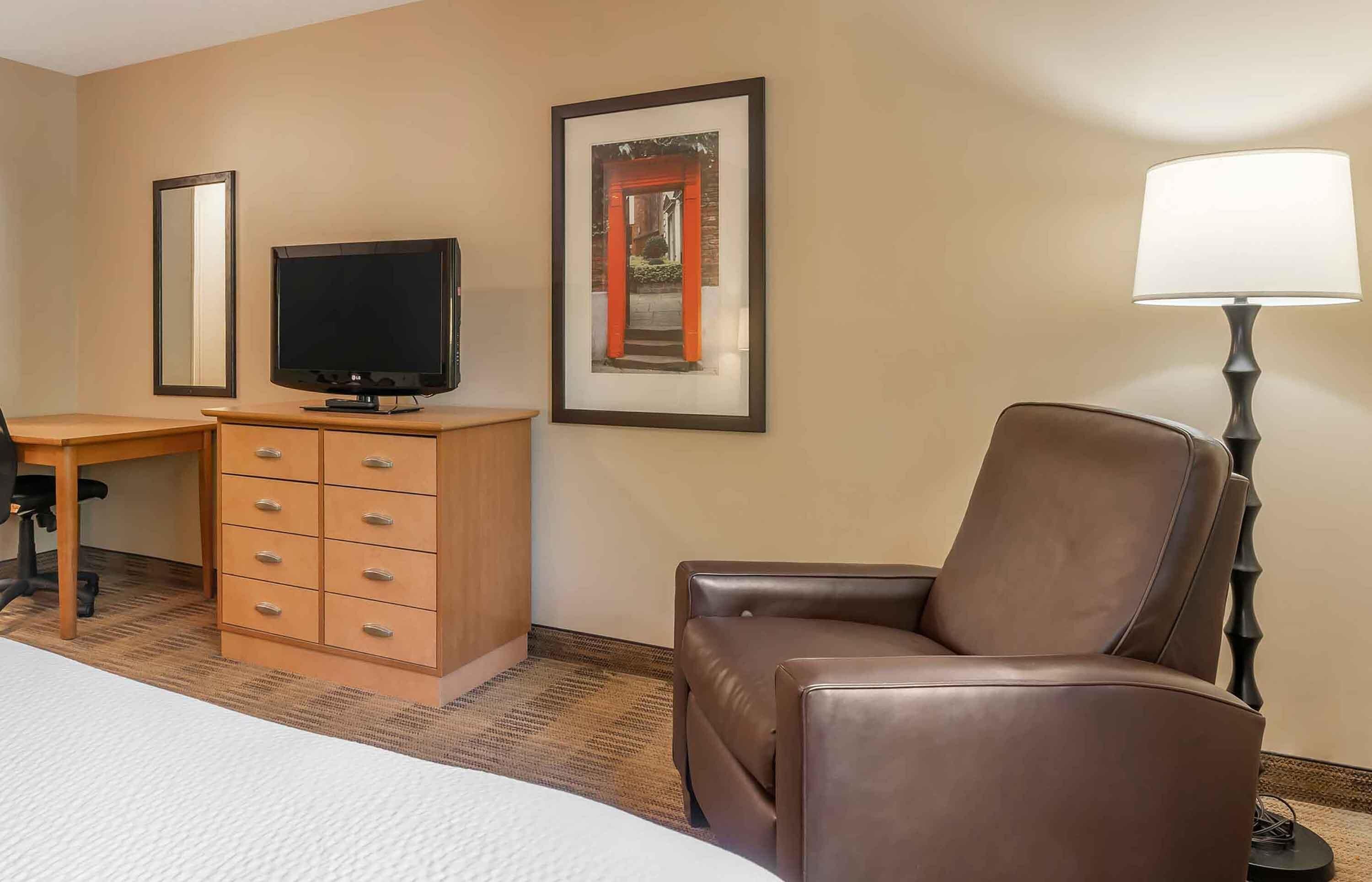 Quarto de hotel com cama, escrivaninha com televisão, poltrona de couro marrom, luminária e duas imagens emolduradas na parede.