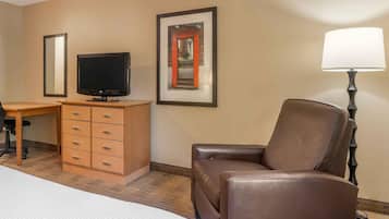 Quarto de hotel com cama, escrivaninha com televisão, poltrona de couro marrom, luminária e duas imagens emolduradas na parede.
