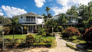 Uma casa de dois andares com varanda, rodeada por um jardim bem cuidado com diversas plantas e um caminho que leva à entrada.