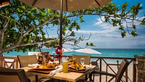 Área de refeições ao ar livre com vista para o mar, com mesa posta, comida e bebidas, rodeada por cadeiras e guarda-sóis.