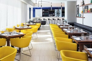 Restaurante com cadeiras amarelas, mesas de madeira e um bar ao fundo.