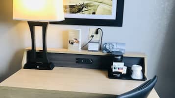 Quarto de hotel com escrivaninha, uma cadeira preta, luminária, quadro emoldurado, cafeteira e pequena mesa com uma xícara e sachê.