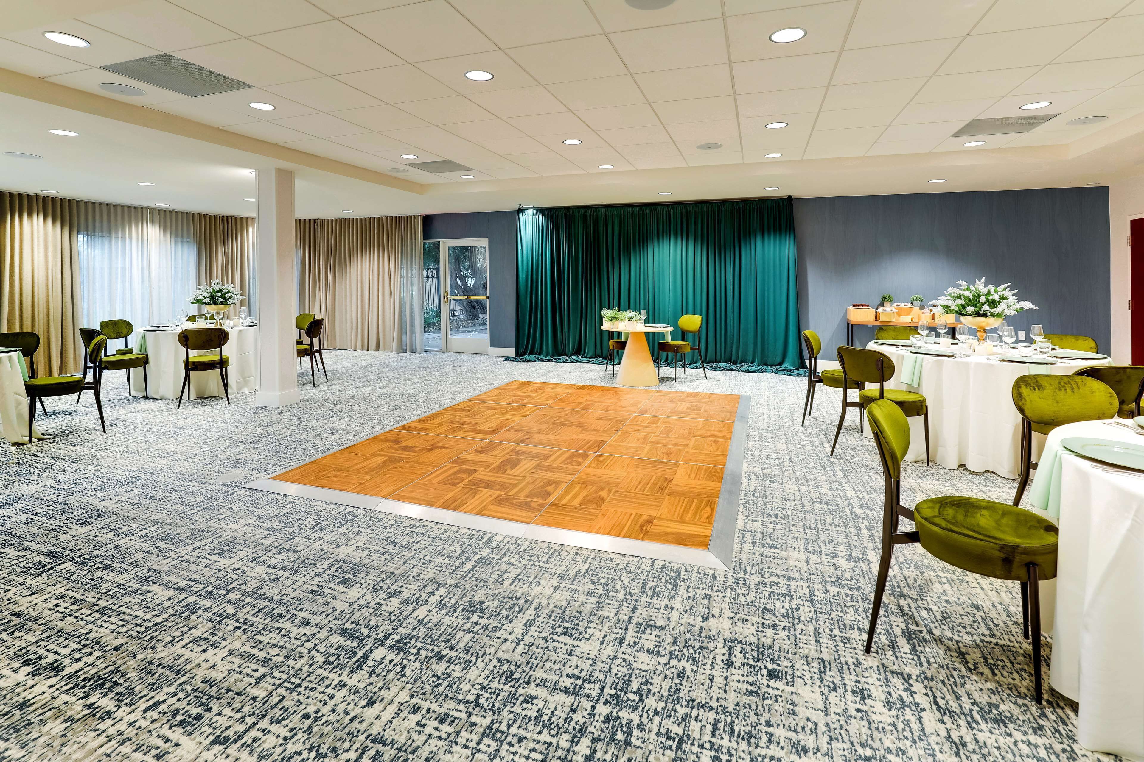 Uma sala de conferências ampla, com um grande piso de madeira, várias mesas preparadas para refeições e cadeiras verdes.