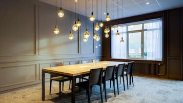 Uma sala de conferências com uma mesa comprida, várias cadeiras, luminárias pendentes, uma janela grande com cortinas e um radiador.