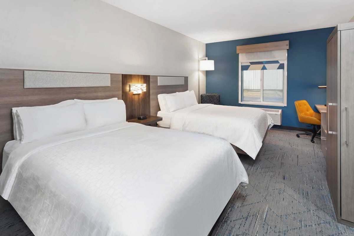 Quarto de hotel com duas camas, uma escrivaninha, uma cadeira, um abajur e janela com vista.
