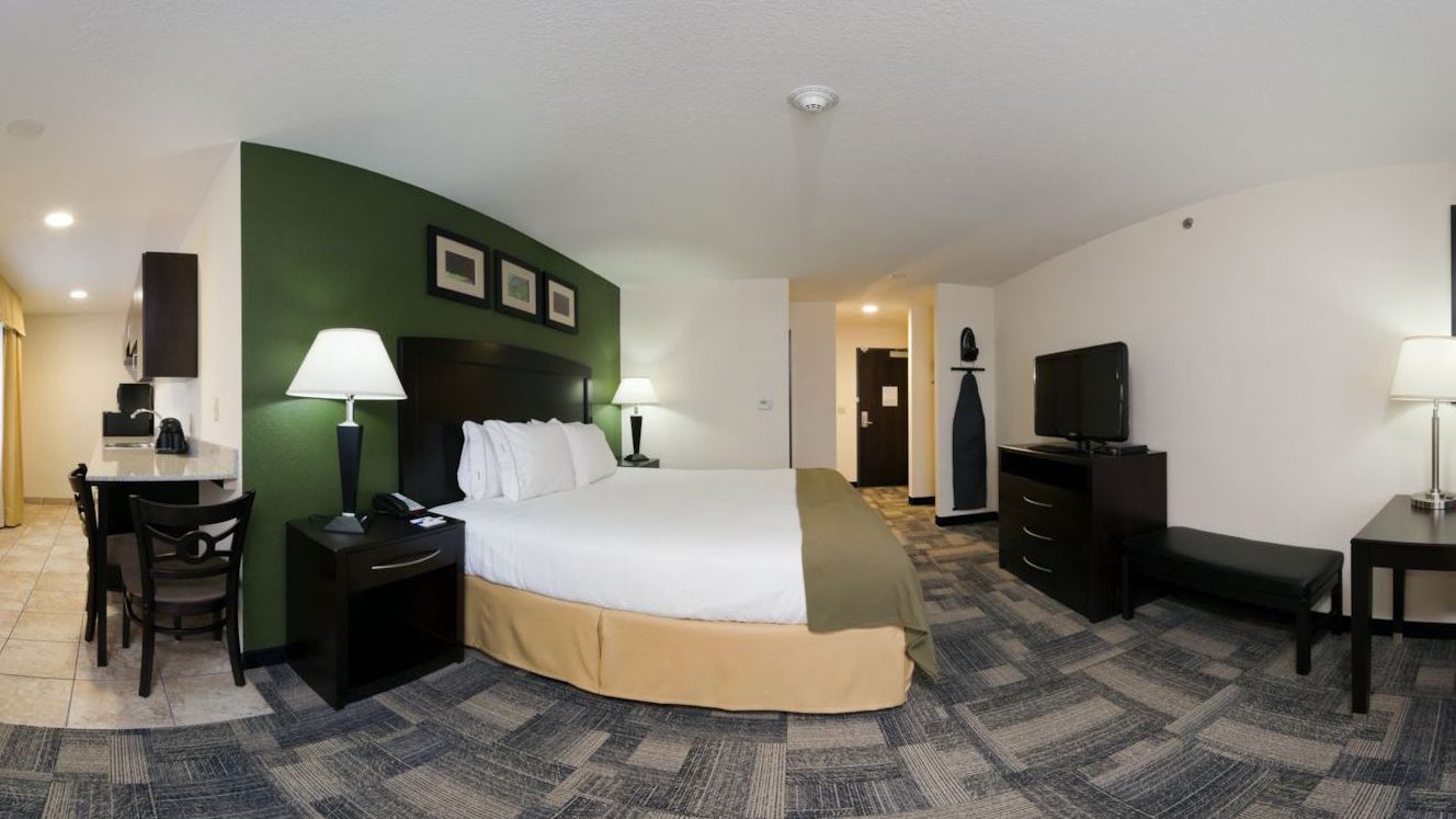 Quarto de hotel com cama, mesas de cabeceira, escrivaninha, cadeira, televisão e luminária.