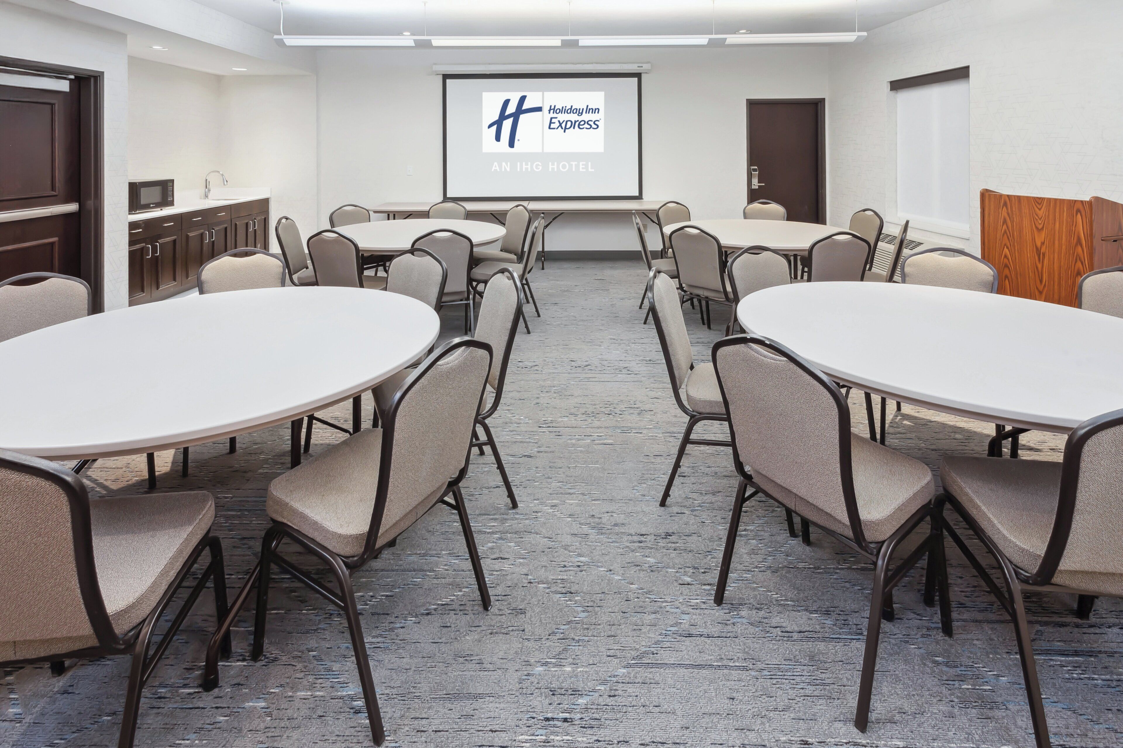 Uma sala de conferências com mesas e cadeiras redondas, uma tela de projeção exibindo o logotipo do Holiday Inn Express e uma área de cozinha compacta.