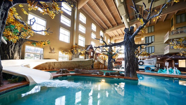 Uma piscina interior com efeito cascata, rodeada por árvores artificiais e uma estrutura com telhado de palha.