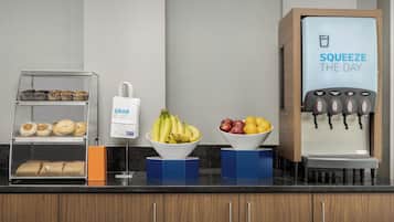 Uma mesa de pequeno-almoço com um saco de lembranças, uma taça de bananas, uma taça de maçãs e laranjas, e um dispensador de bebidas.