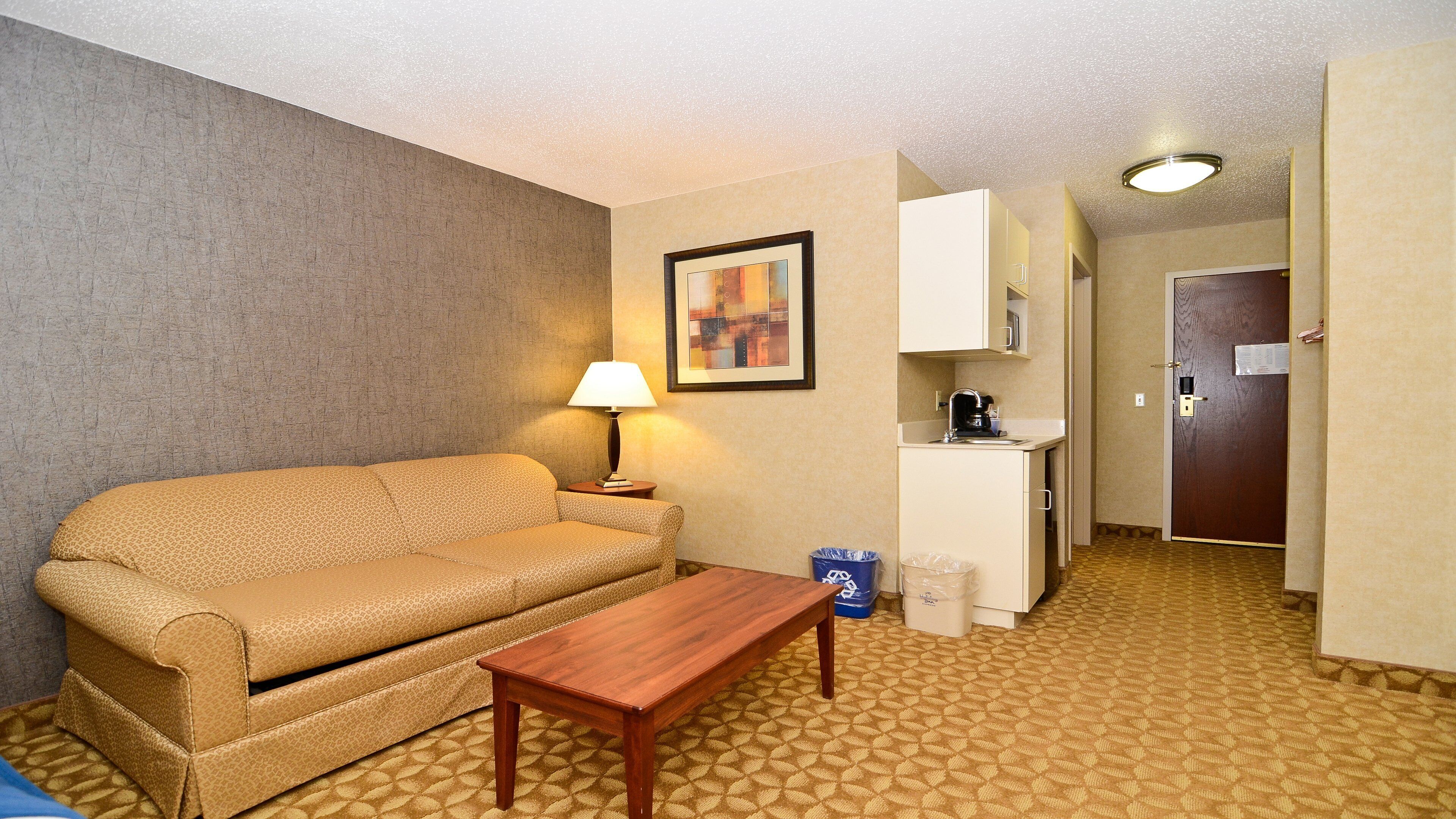 Quarto de hotel com sofá, mesa pequena, luminária, cozinha compacta e porta de acesso a outro cômodo.