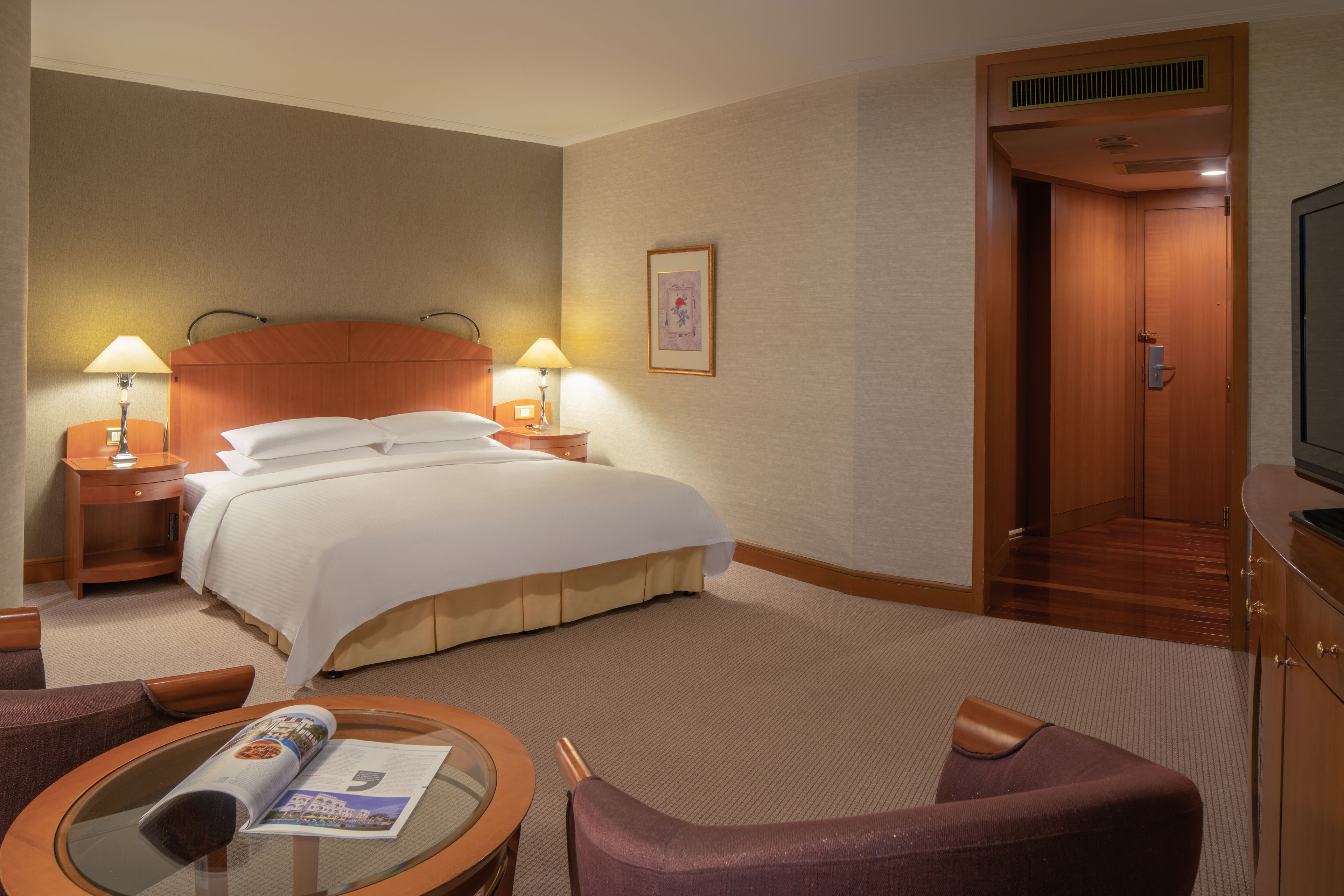 Quarto de hotel com uma cama grande, duas luminárias de cabeceira, um painel de cabeceira de madeira, um quadro na parede, um armário de madeira e uma pequena mesa com uma revista.