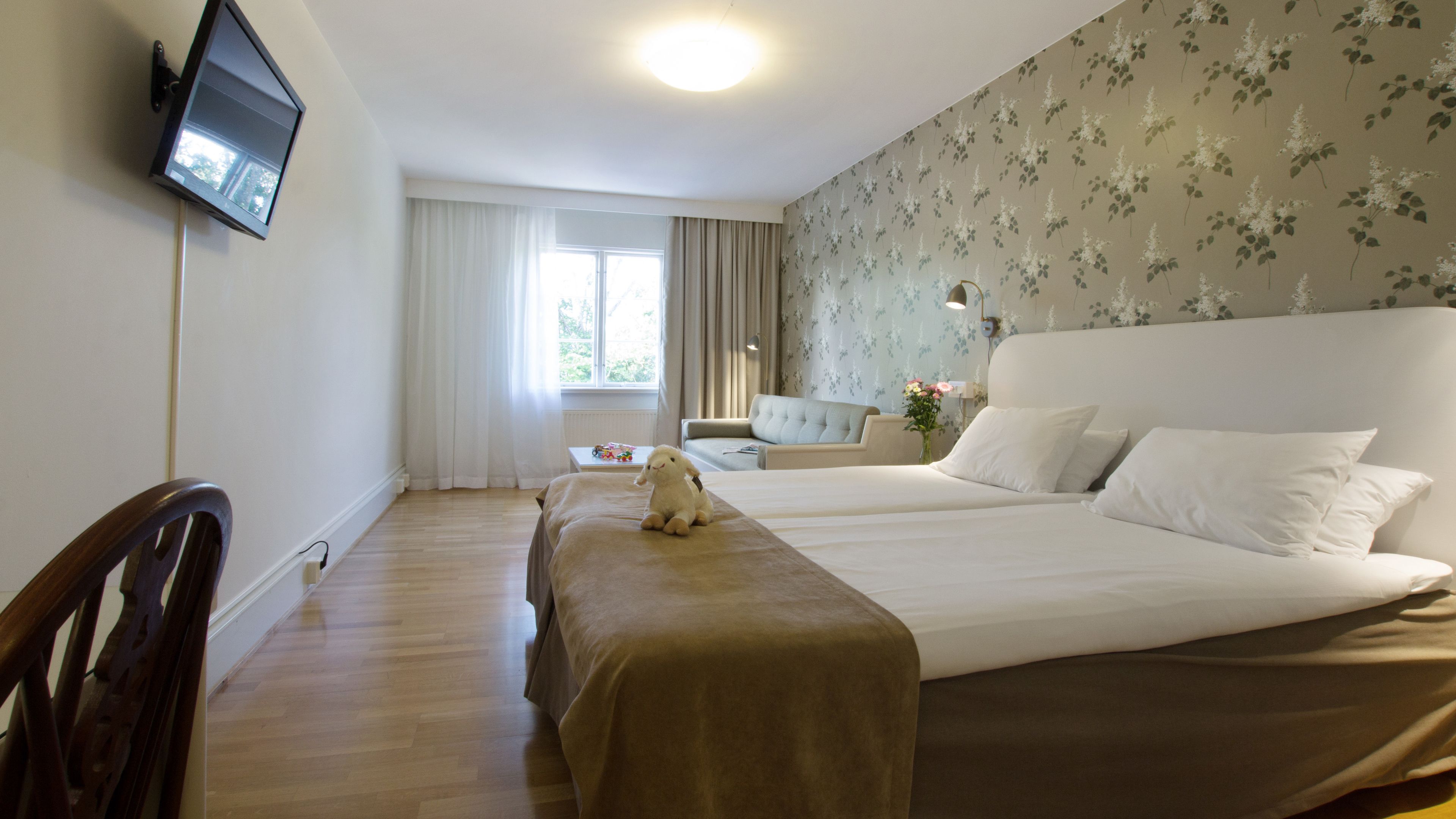 Quarto de hotel com duas camas, uma TV fixada na parede, uma janela com cortinas e papel de parede floral.