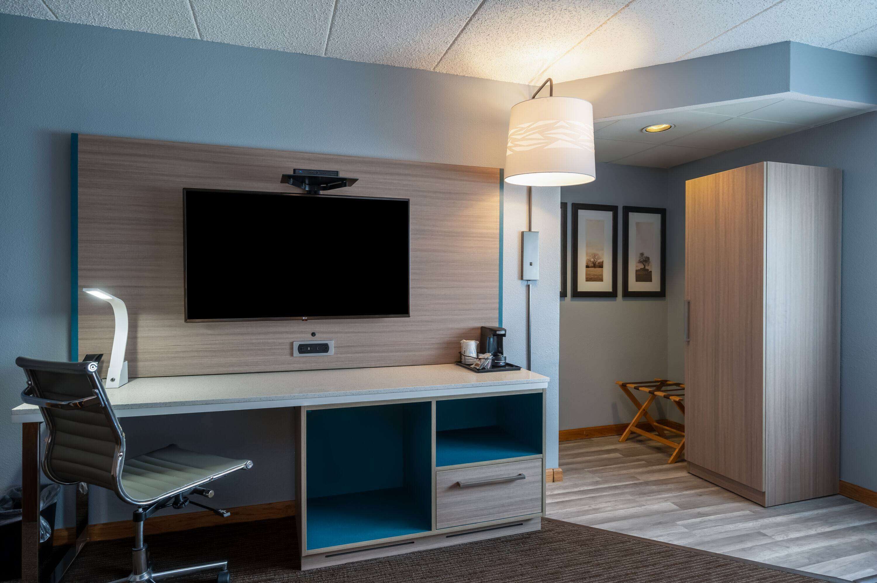 Quarto de hotel moderno com TV de tela plana fixada numa parede de madeira, uma escrivaninha com cadeira, uma luminária e um guarda-roupa.