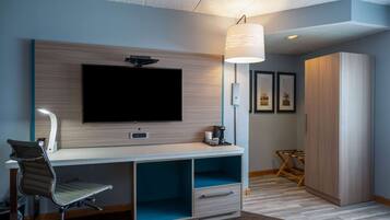 Quarto de hotel moderno com TV de tela plana fixada numa parede de madeira, uma escrivaninha com cadeira, uma luminária e um guarda-roupa.