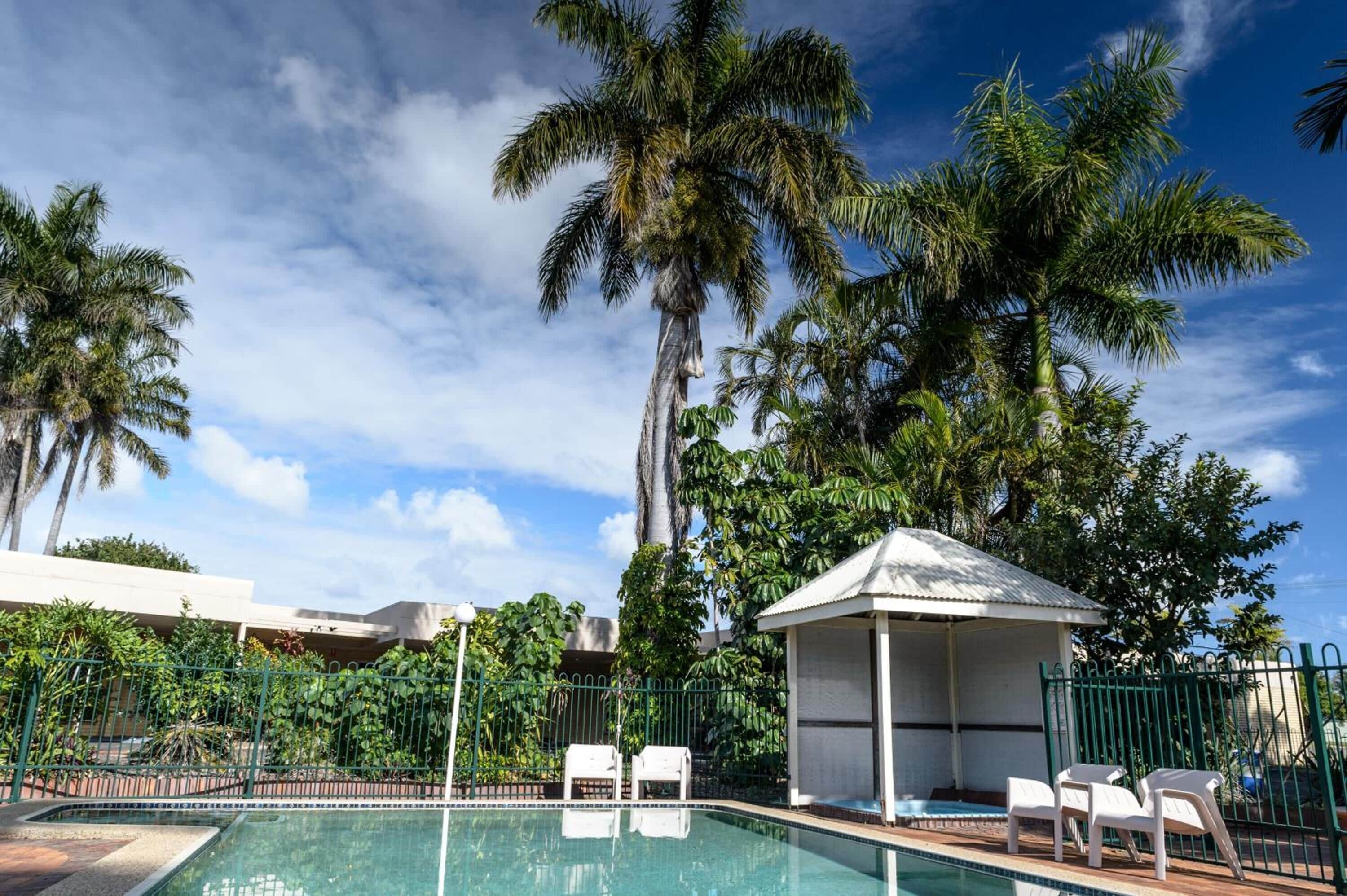 Uma piscina com espreguiçadeiras e um gazebo, cercada por palmeiras e um céu claro.