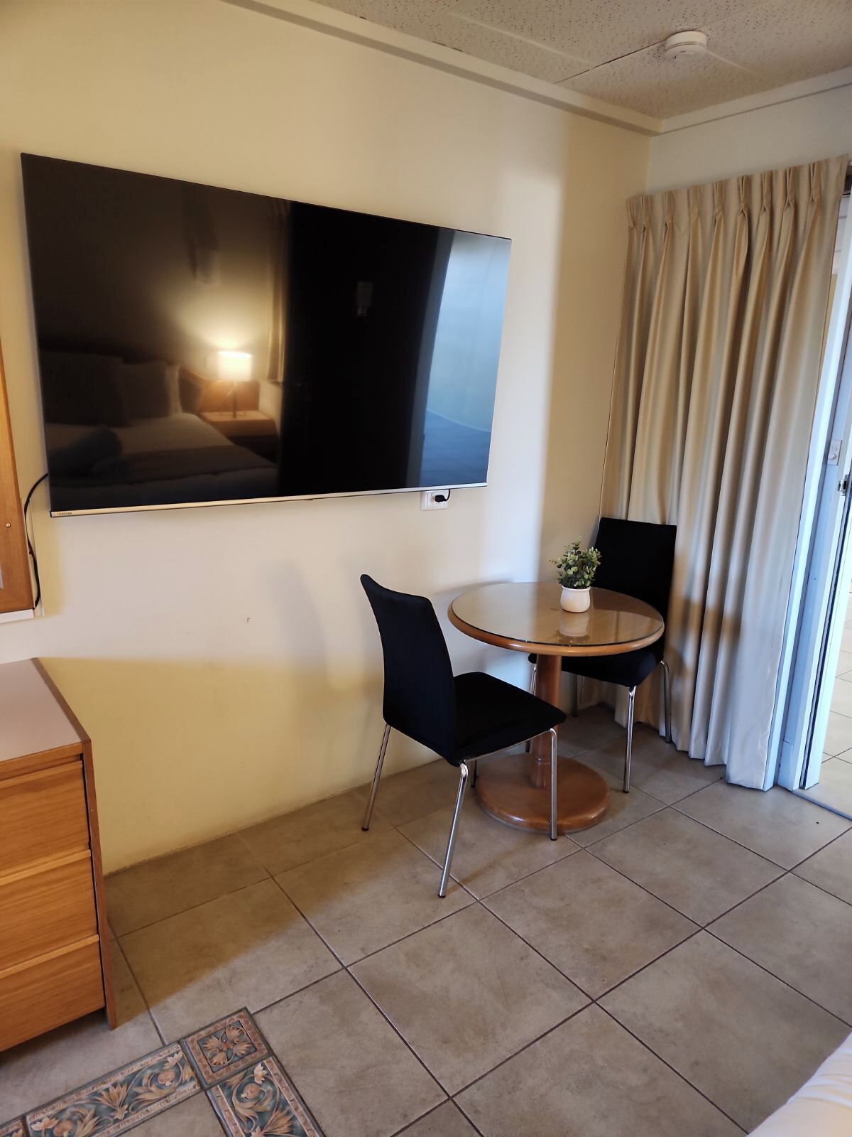 Quarto de hotel com televisão de tela plana na parede, uma mesa de jantar redonda com duas cadeiras, uma pequena planta em vaso e um armário de madeira.