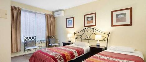 Quarto de hotel com duas camas, uma mesinha, uma cadeira e uma janela com cortinas.