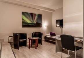 Quarto de hotel moderno com uma grande pintura da aurora boreal, uma cama com travesseiro roxo, uma escrivaninha com computador e duas poltronas de couro.