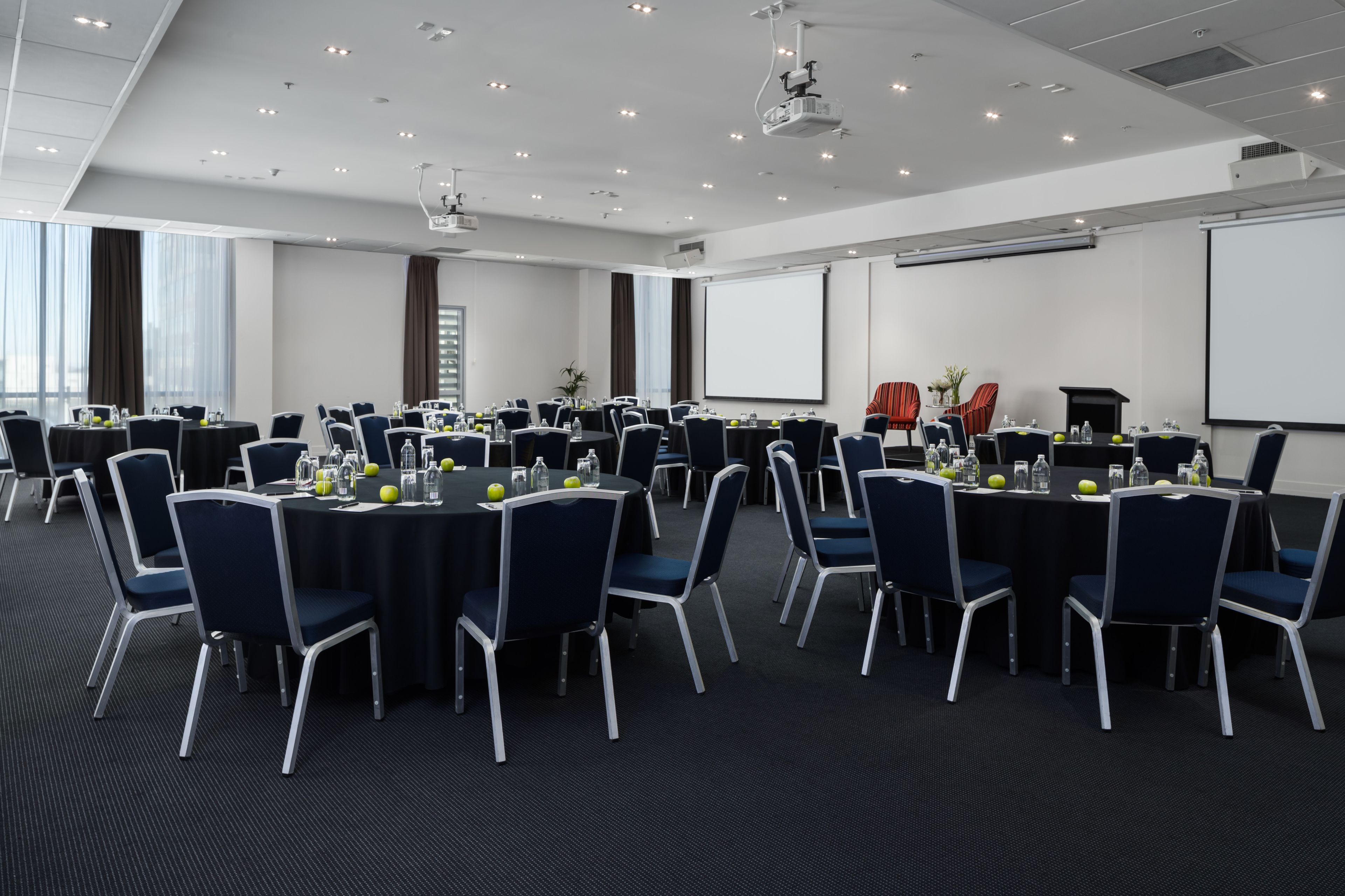 Sala de conferência com mesas redondas, cadeiras e tela de projetor.