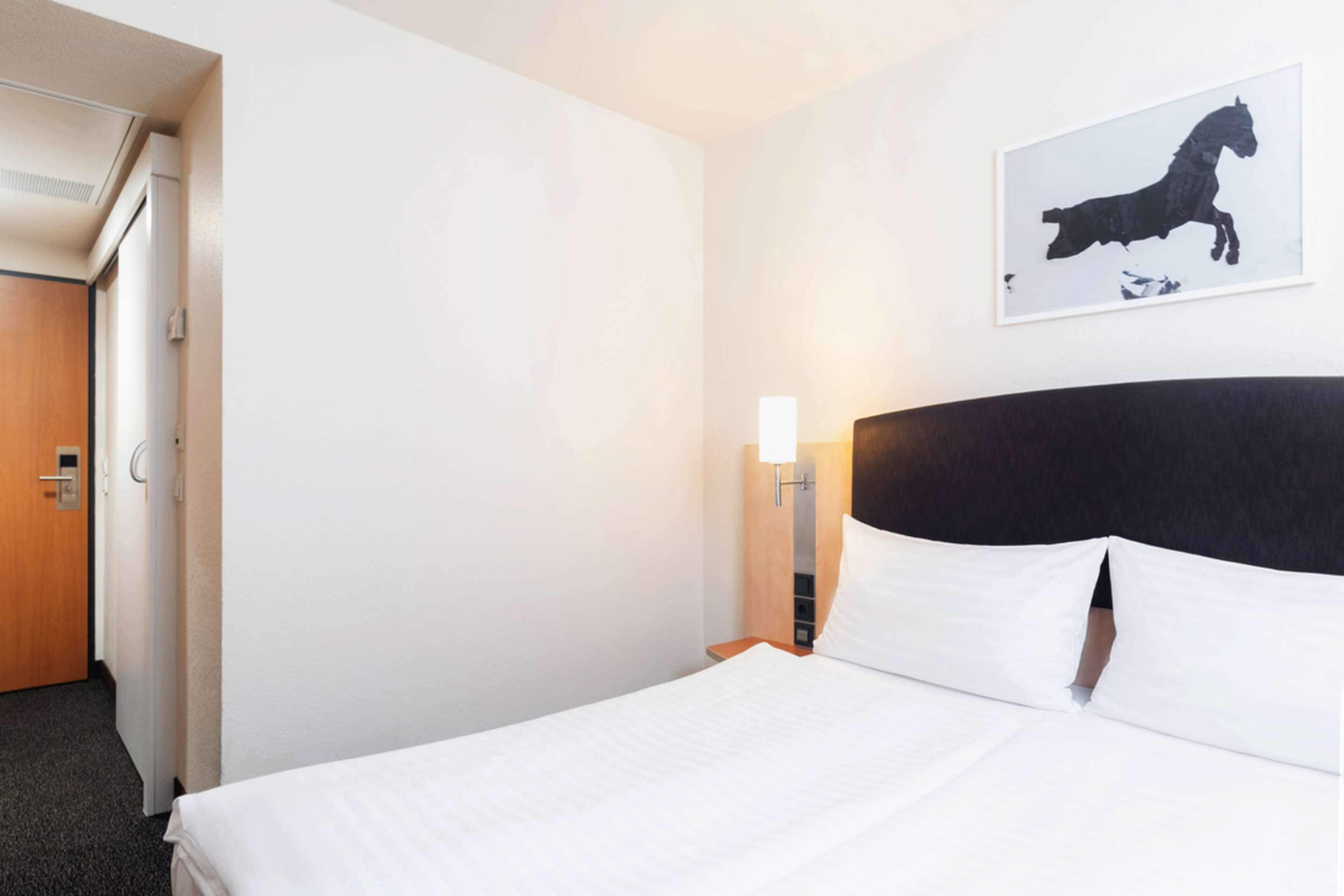 Quarto de hotel com cama, mesa de cabeceira, luminária na parede e uma porta.