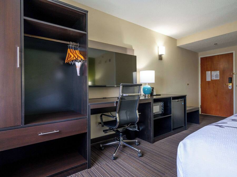 Quarto de hotel com cama, escrivaninha, computador, televisão, guarda-roupa e abajur.