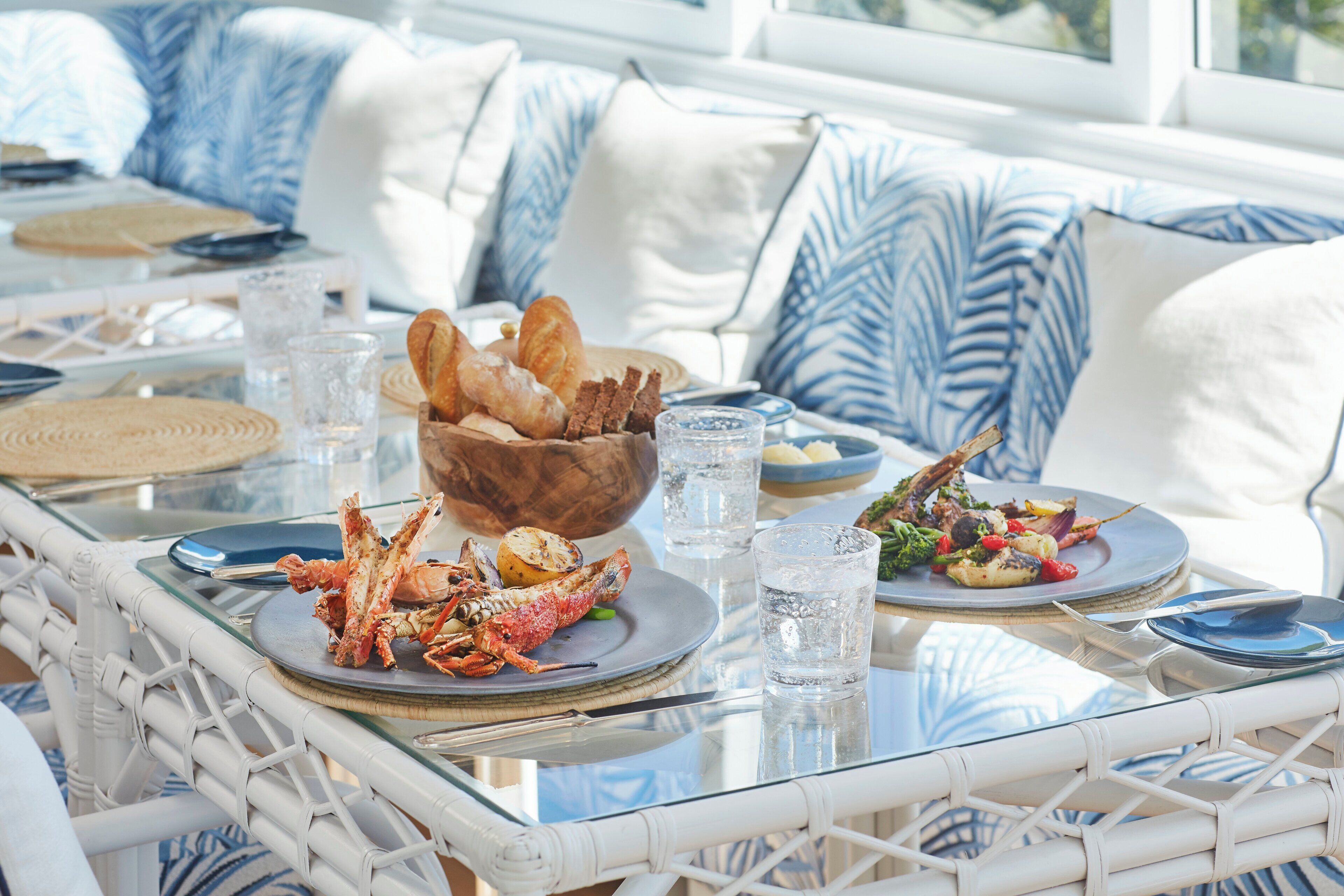 Mesa de jantar com pratos de frutos do mar, pão e bebidas, num espaço iluminado com mobiliário de vime branco e almofadas estampadas em azul.