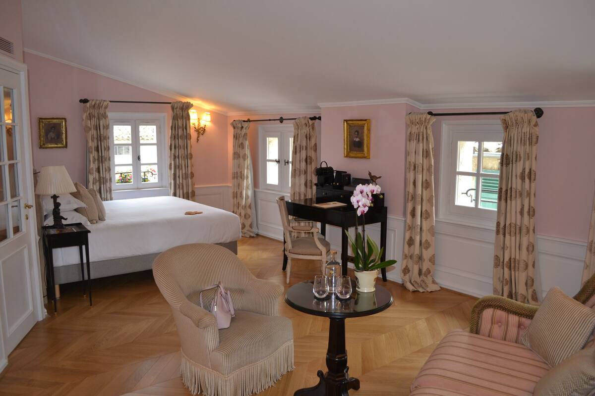 Quarto de hotel com uma cama, duas poltronas, uma mesinha de centro com um vaso de flores, um piano e uma janela com cortinas.