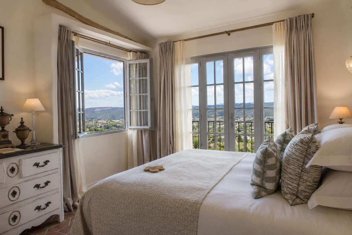 Quarto com uma cama grande, duas janelas com cortinas, cômoda com luminárias e vista para o campo.