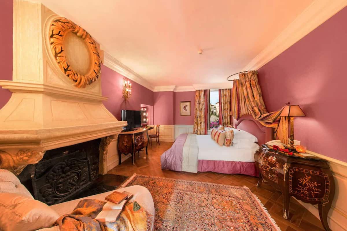 Um quarto com lareira, cama com dossel, escrivaninha com abajur e um espelho grande na parede.