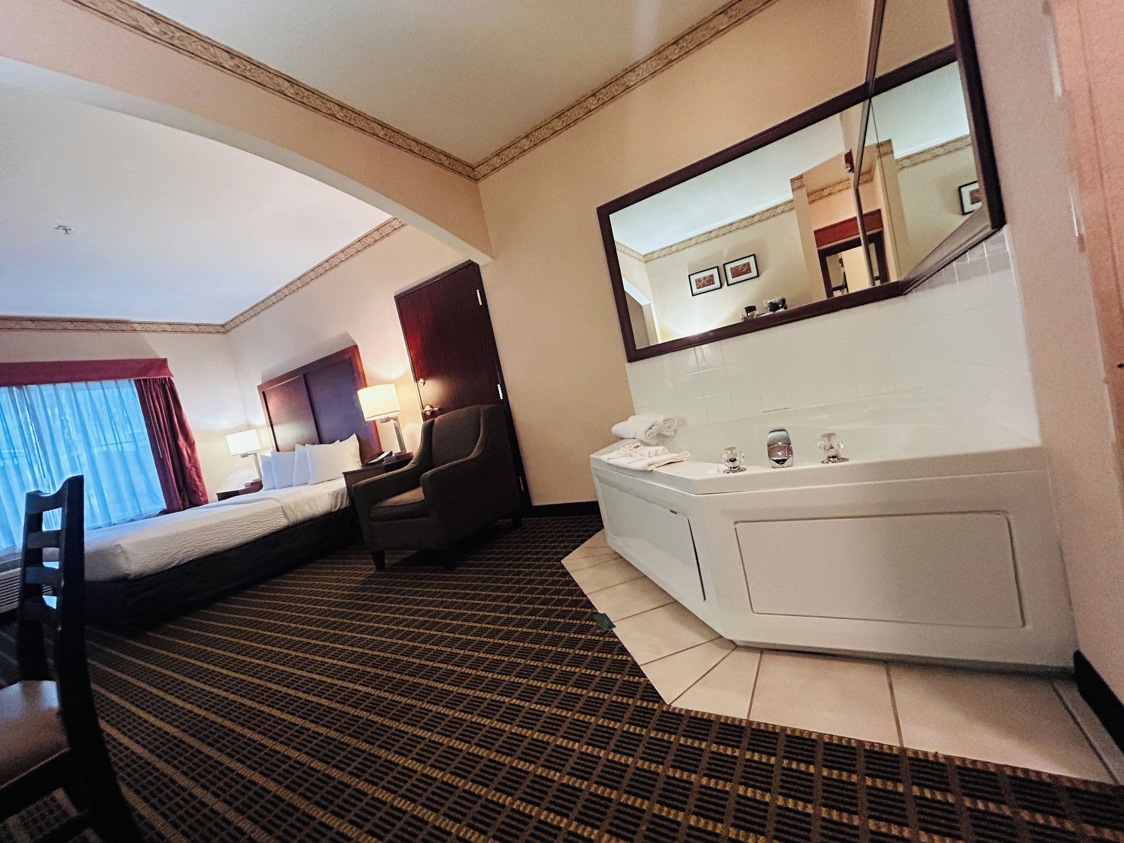 Quarto de hotel com cama, cadeira, escrivaninha, luminária de mesa, espelho e banheiro com banheira e pia.