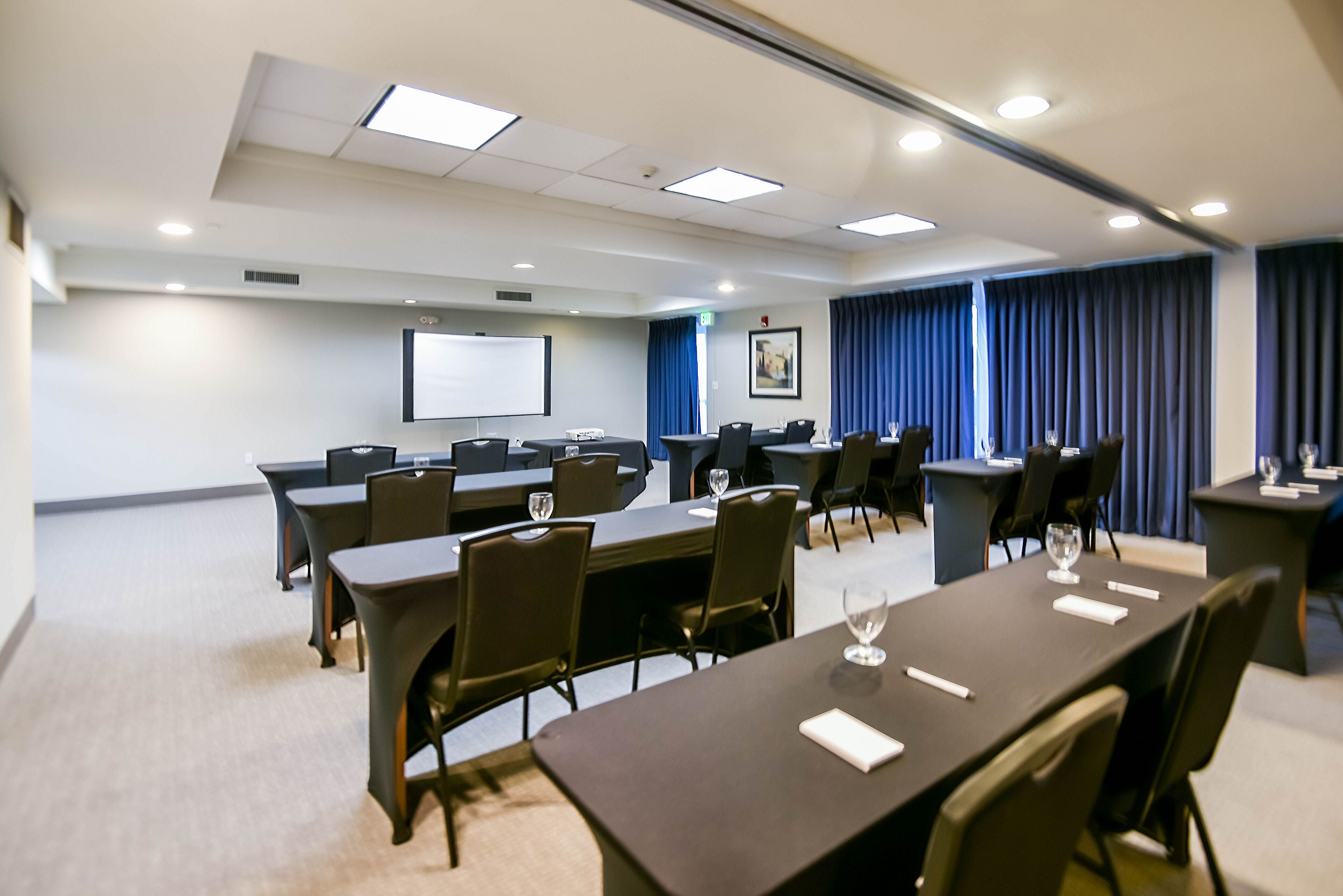 Sala de conferência com várias mesas e cadeiras, uma tela de projetor e uma janela grande com cortinas.